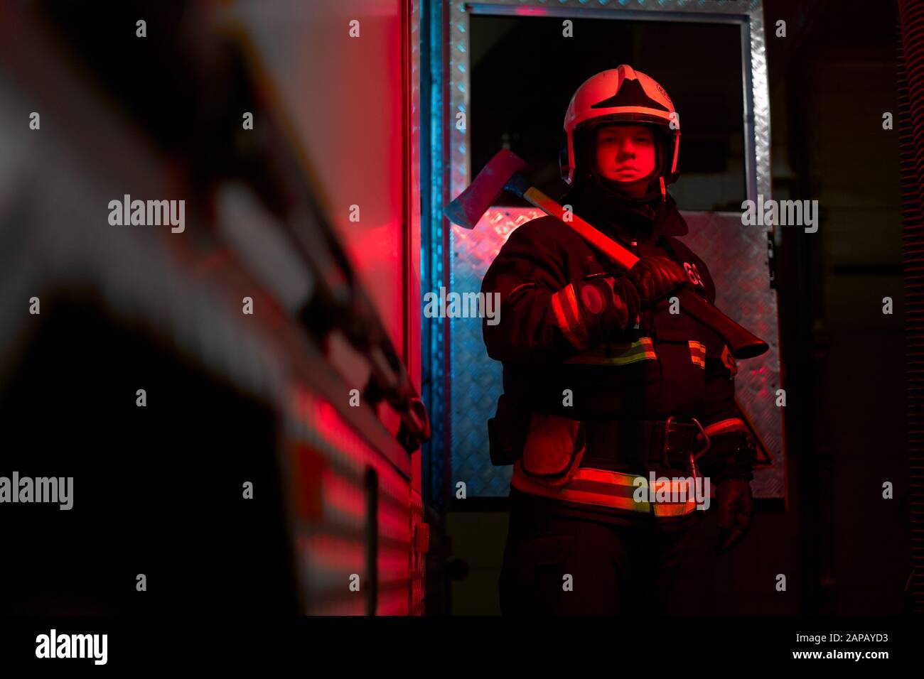Close up firefighter man with hammer on his shoulder standing near fire ...