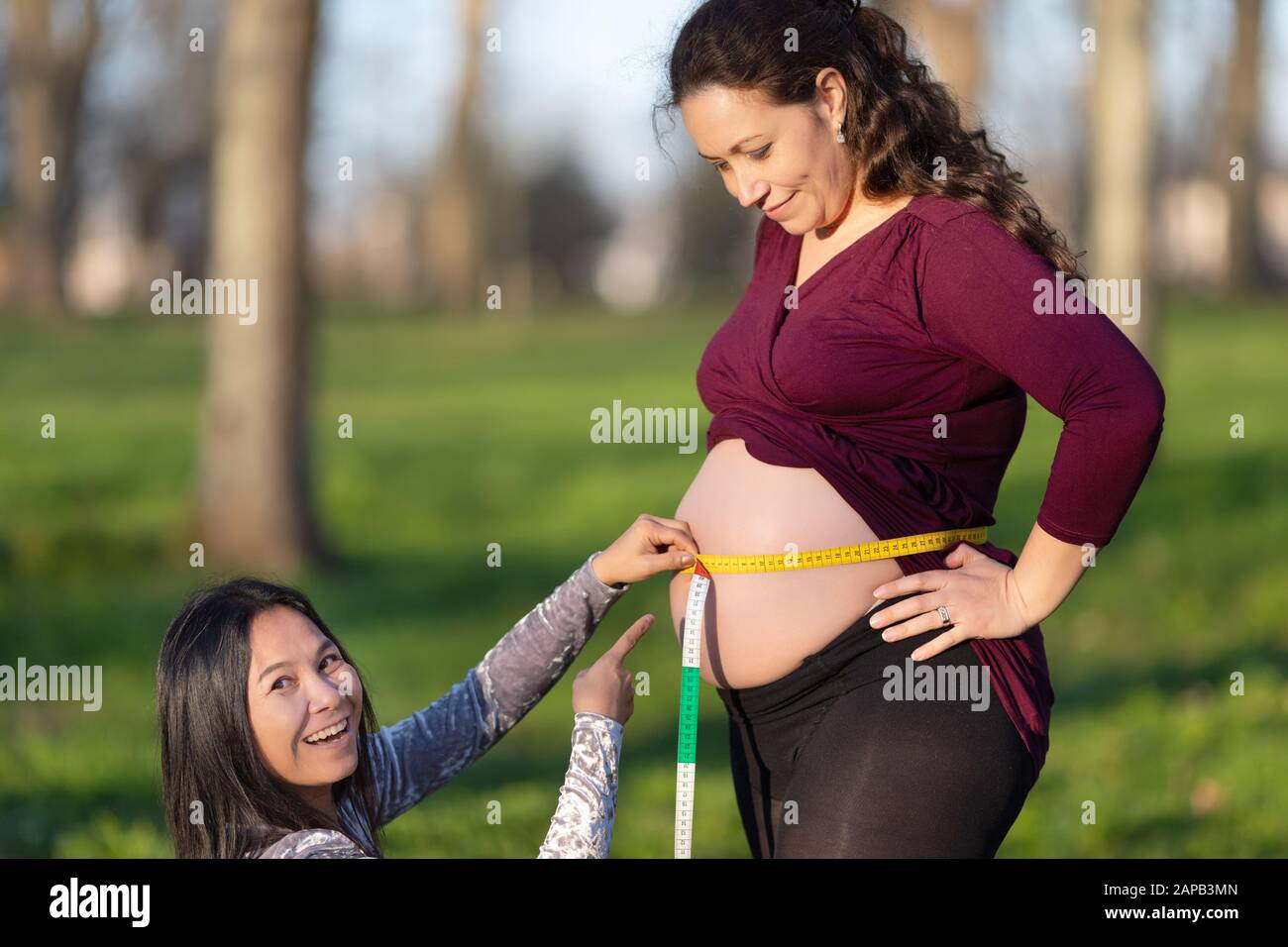 Cheerful Asian woman measuring pregnant womans bare belly with a tape ...