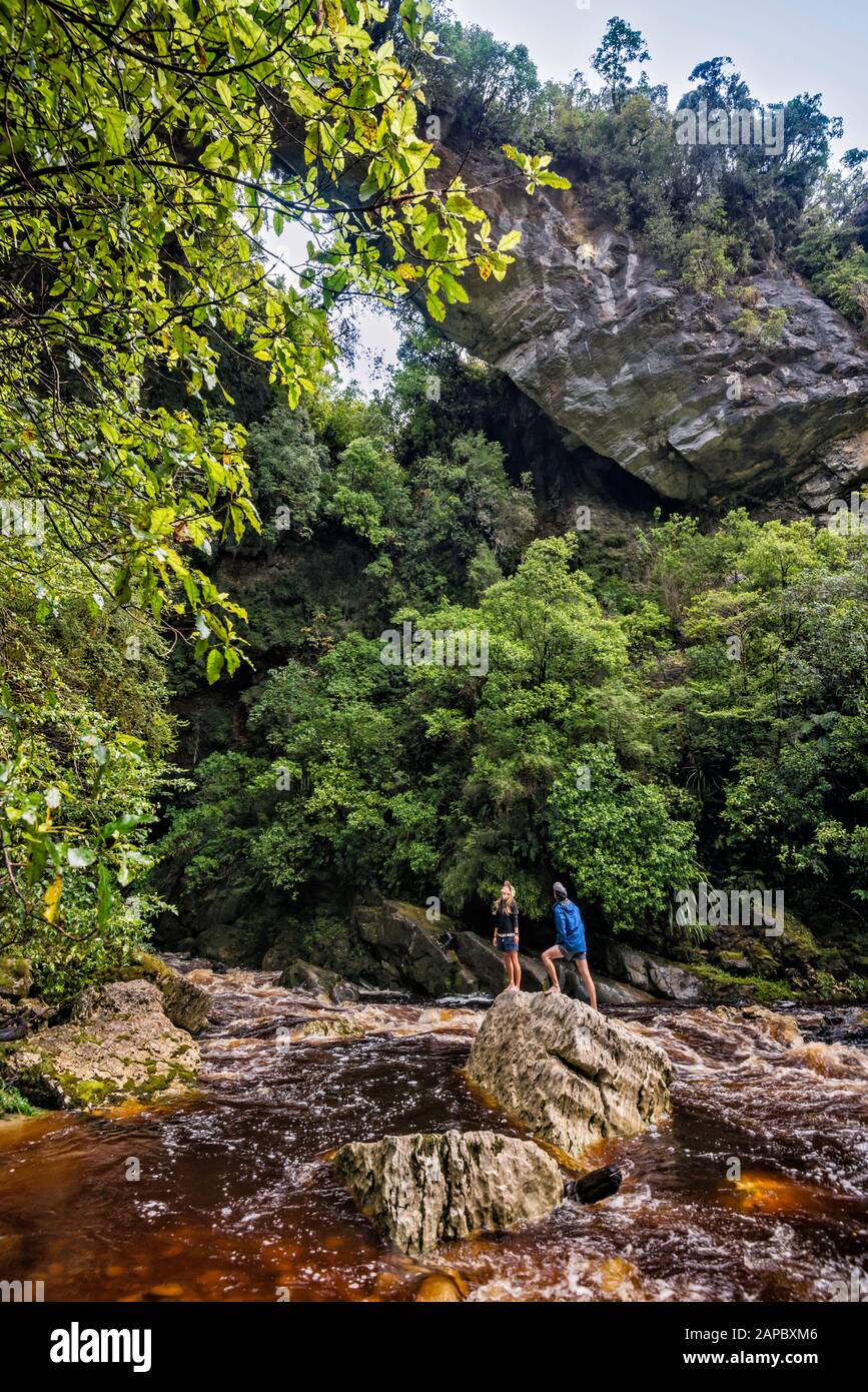 Hikers looking at Oparara Arch, on boulder at Oparara River, rain ...