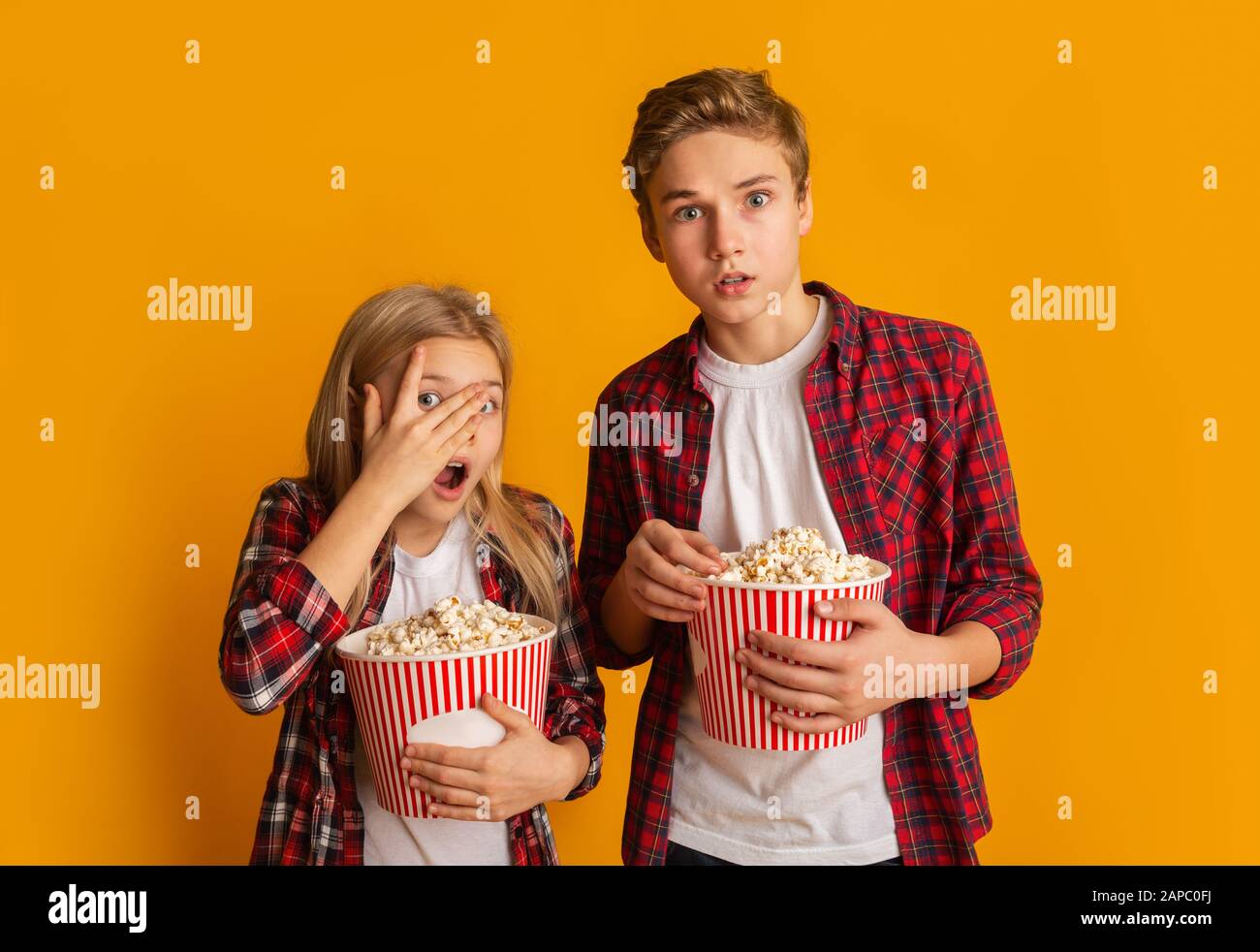 Scared brother and sister holding popcorn buckets and looking at camera ...