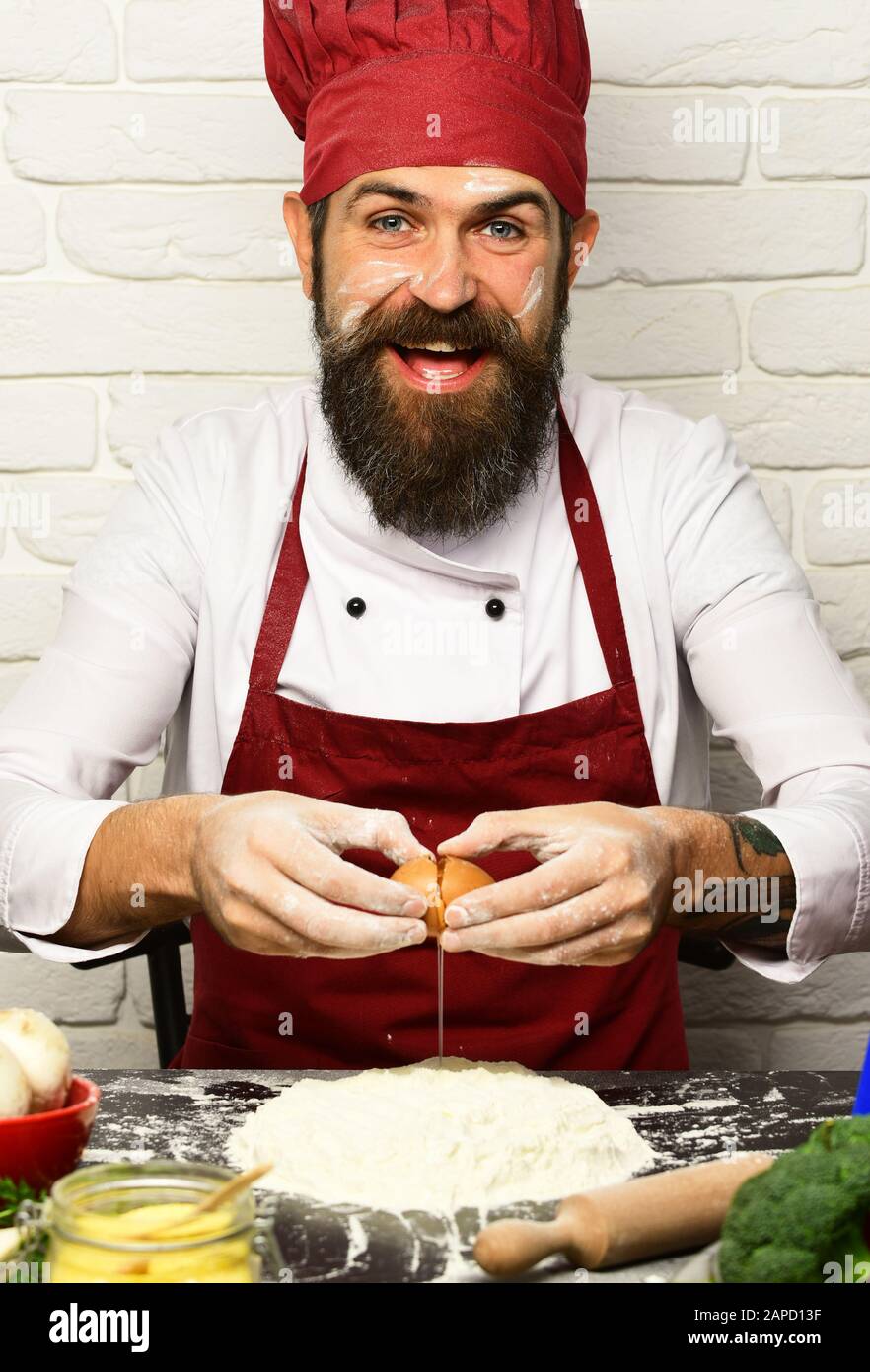 Cook with happy face in burgundy uniform sits by table with flour and ...