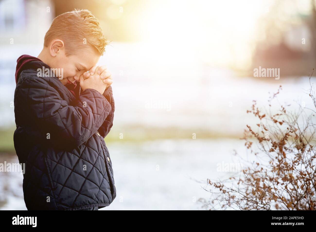 Little boy standing in a park and praying under sunlight with a blurry ...