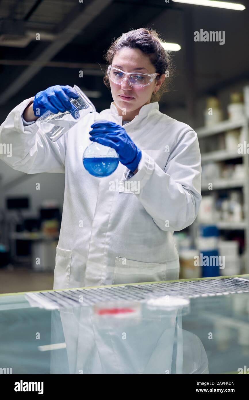 Young lab woman in glasses and white coat with flask with blue liquid ...