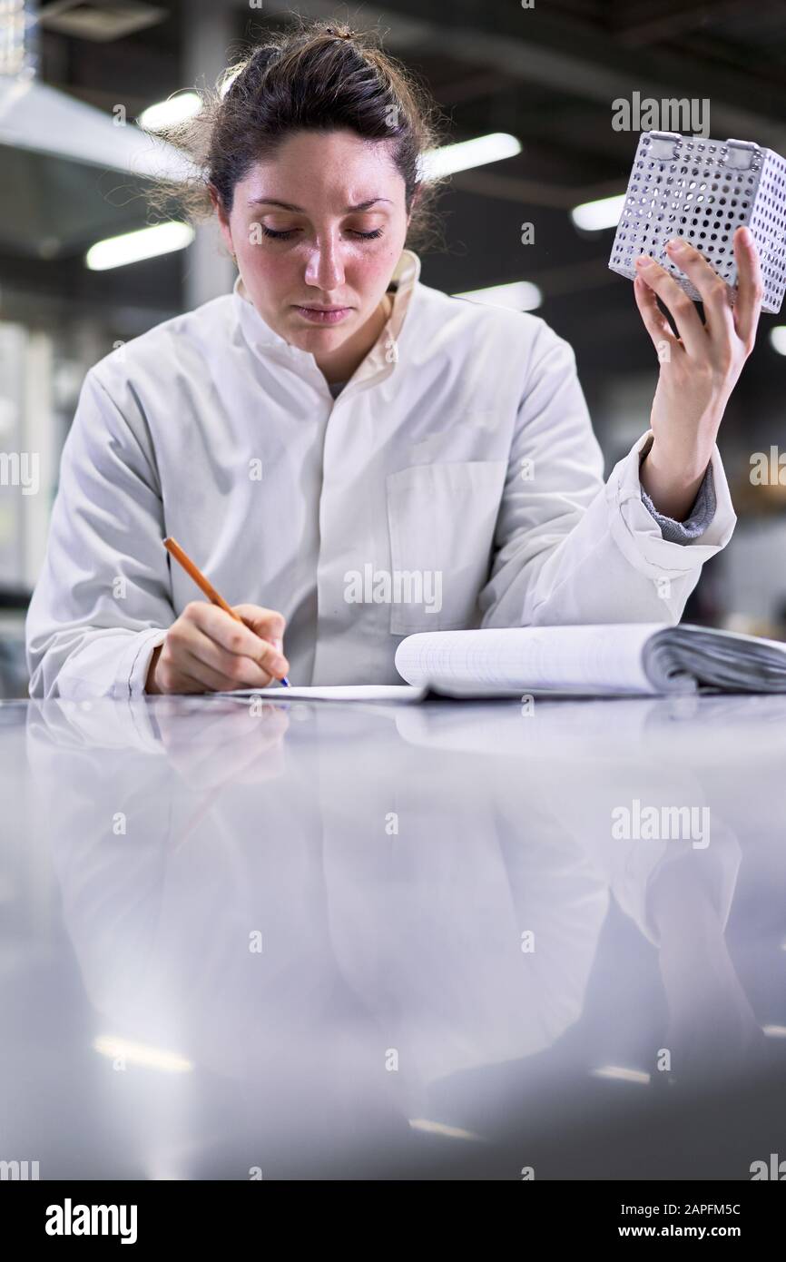 Brunette girl in white coat with metal cube sits on table in laboratory ...