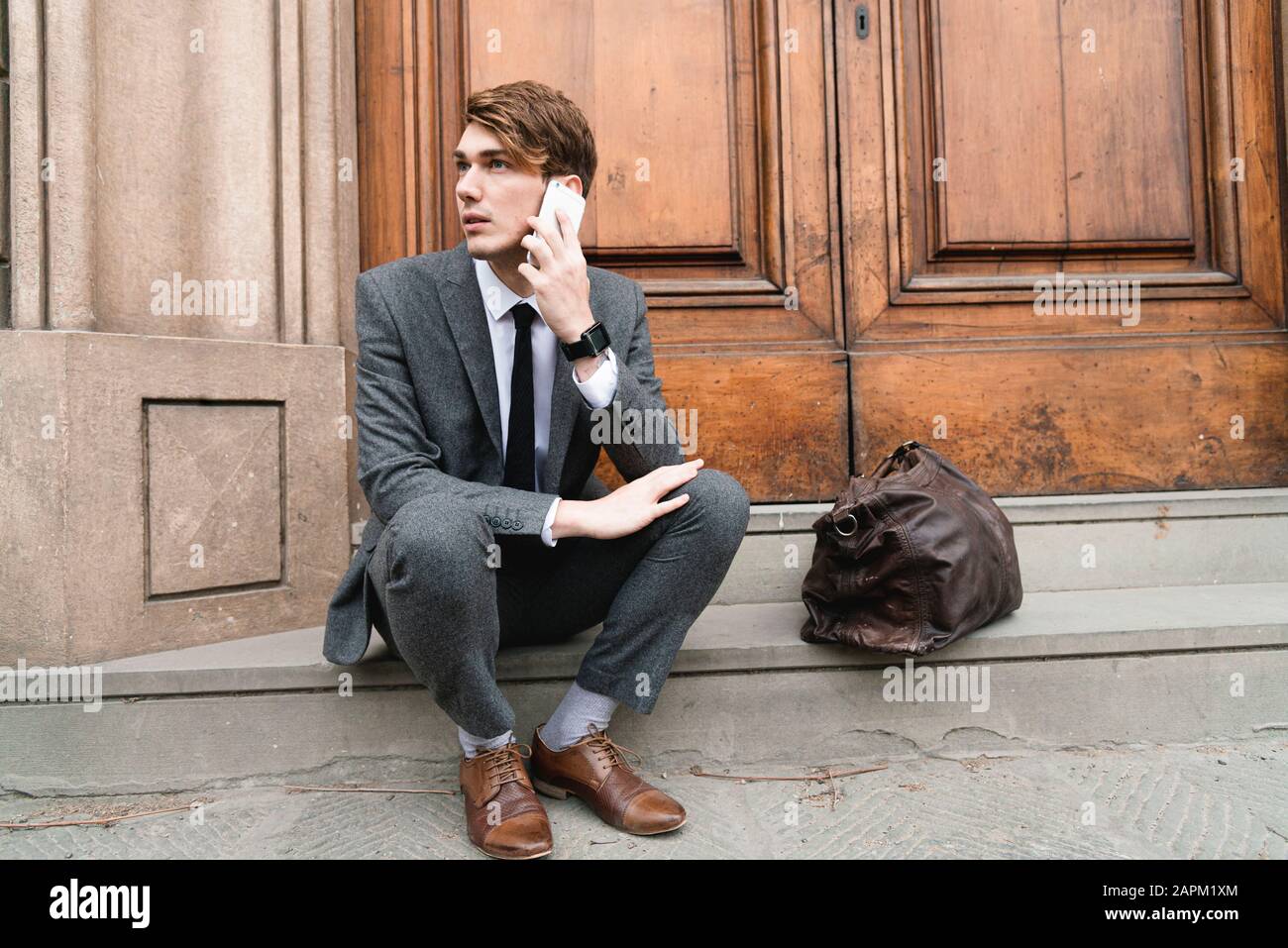 Portrait of young businessman on the phone sitting on steps of house ...