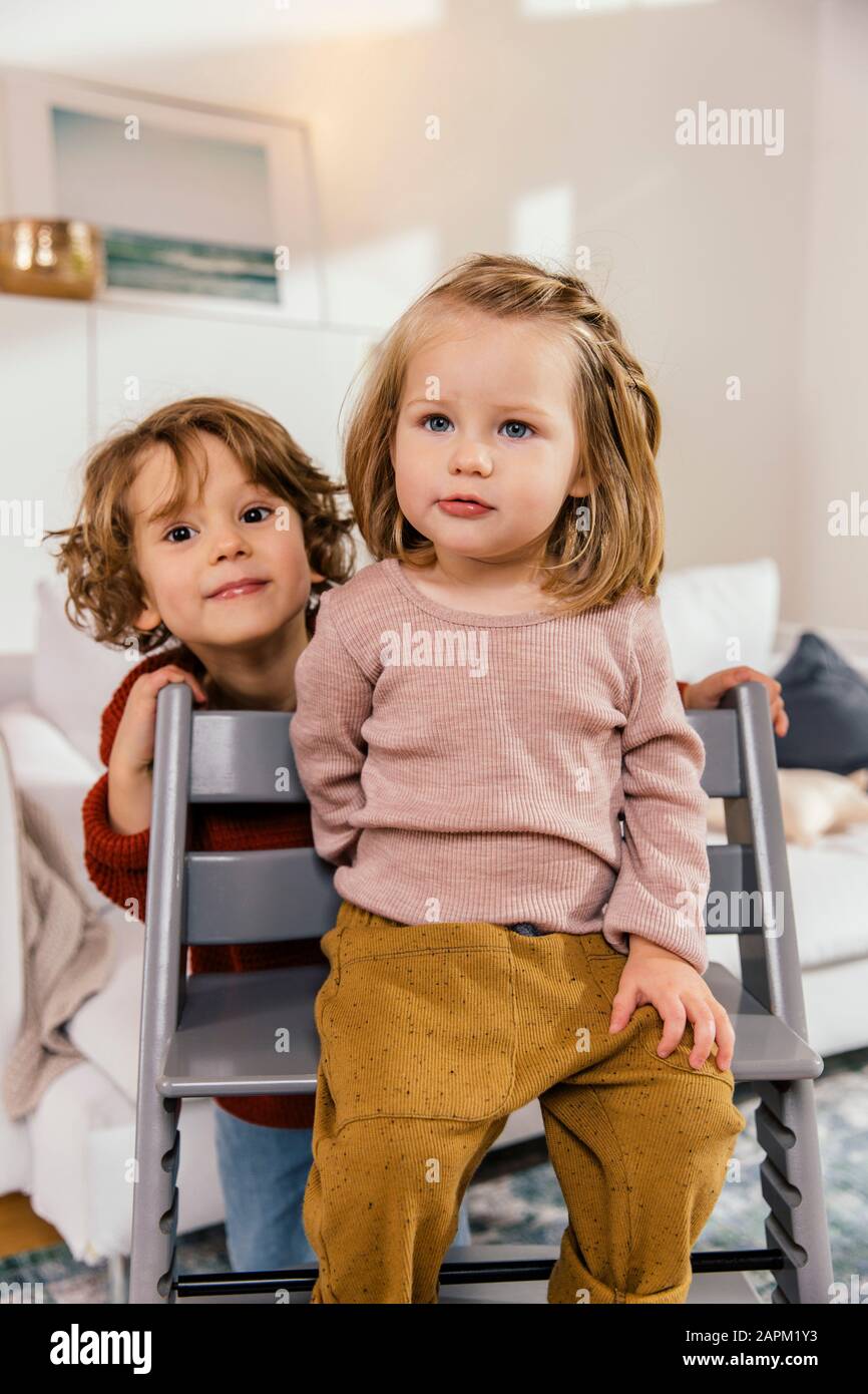 Little girl sitting on high chair at home with sister behind her Stock ...