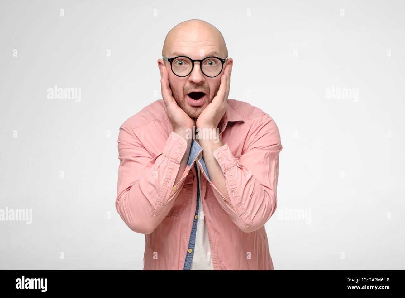Shocked bald man in glasses emotionally holds hands on his face, in ...