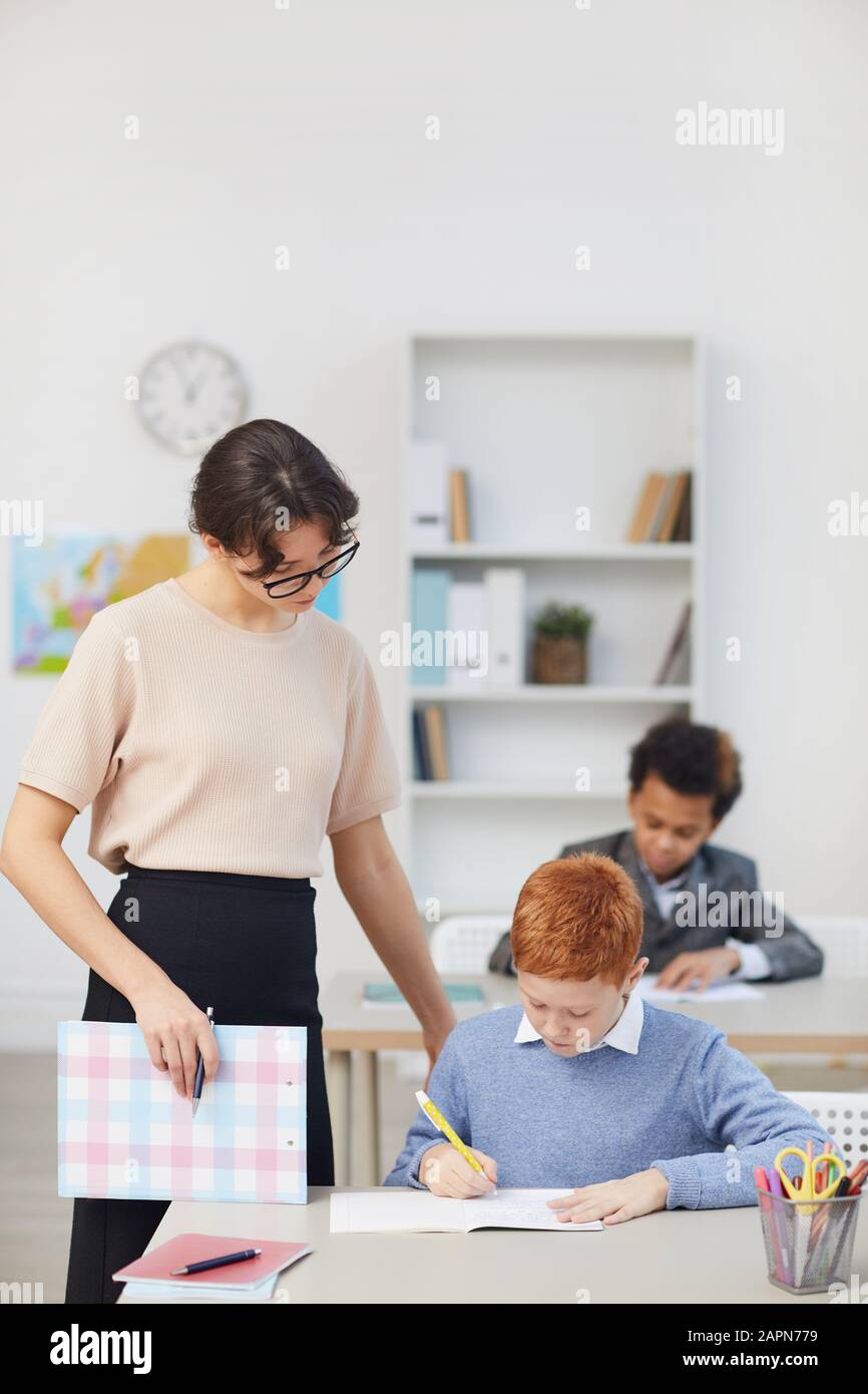 Young female teacher in eyeglasses looking at her student's work while ...