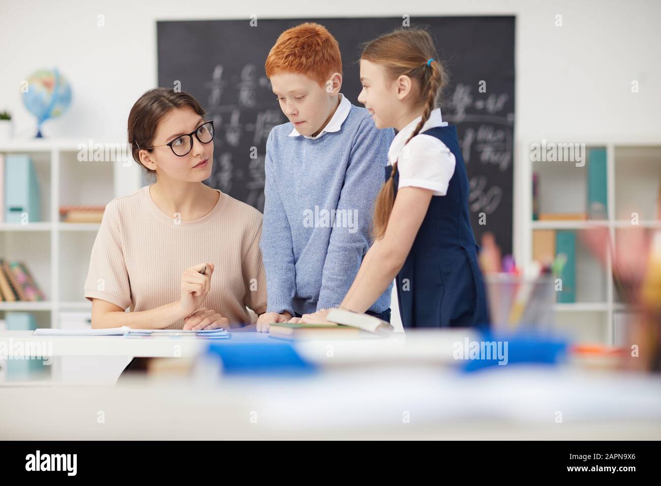 Young teacher in eyeglasses talking to children and giving them the ...