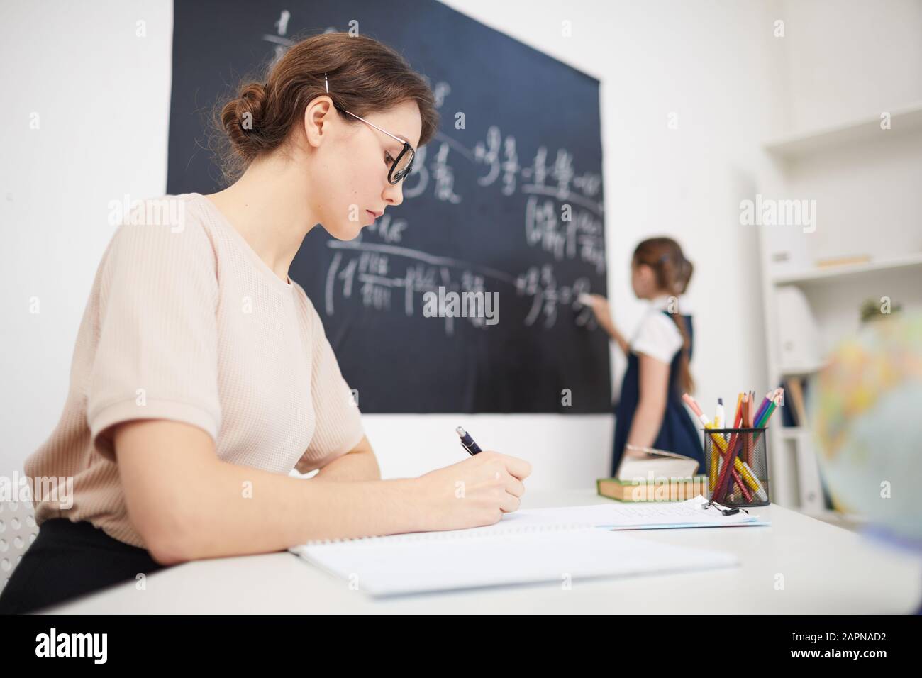 Young teacher sitting at the table and writing while schoolgirl working ...