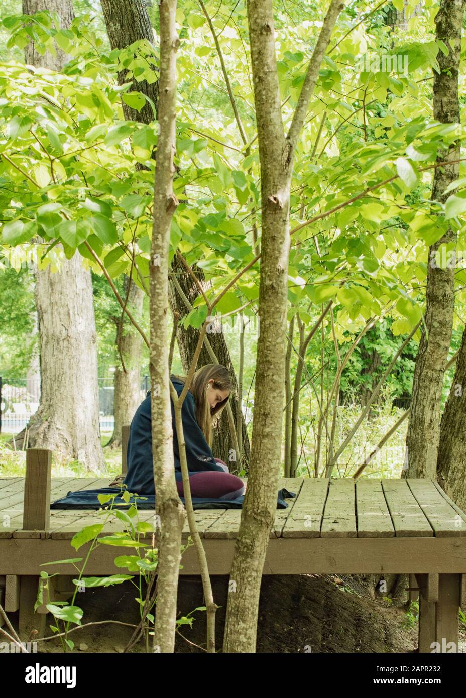 Teenage girl sitting on a deck beneath a stand of trees. She is sitting ...