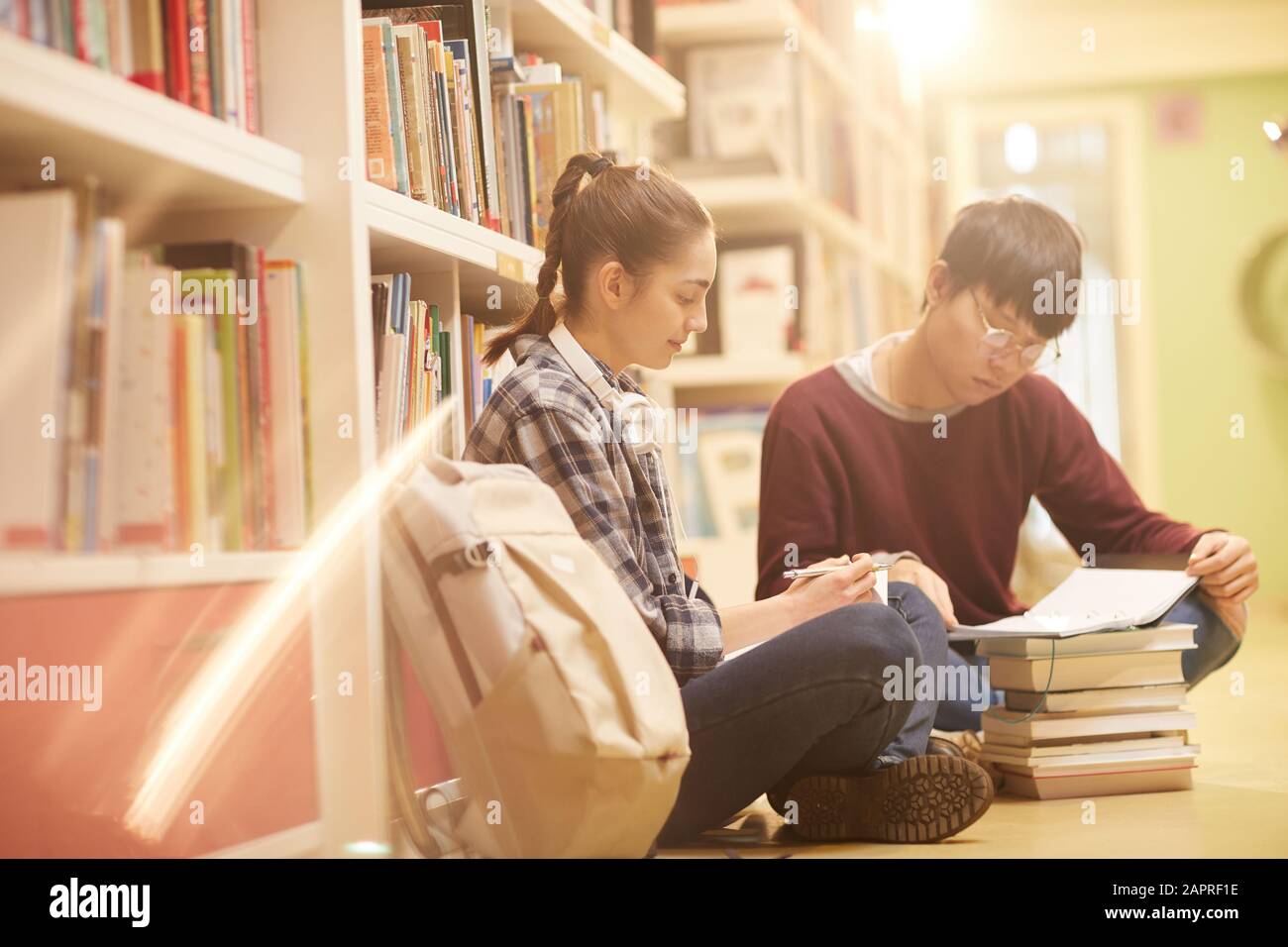 Two students sitting on the floor near the bookcase and reading books ...