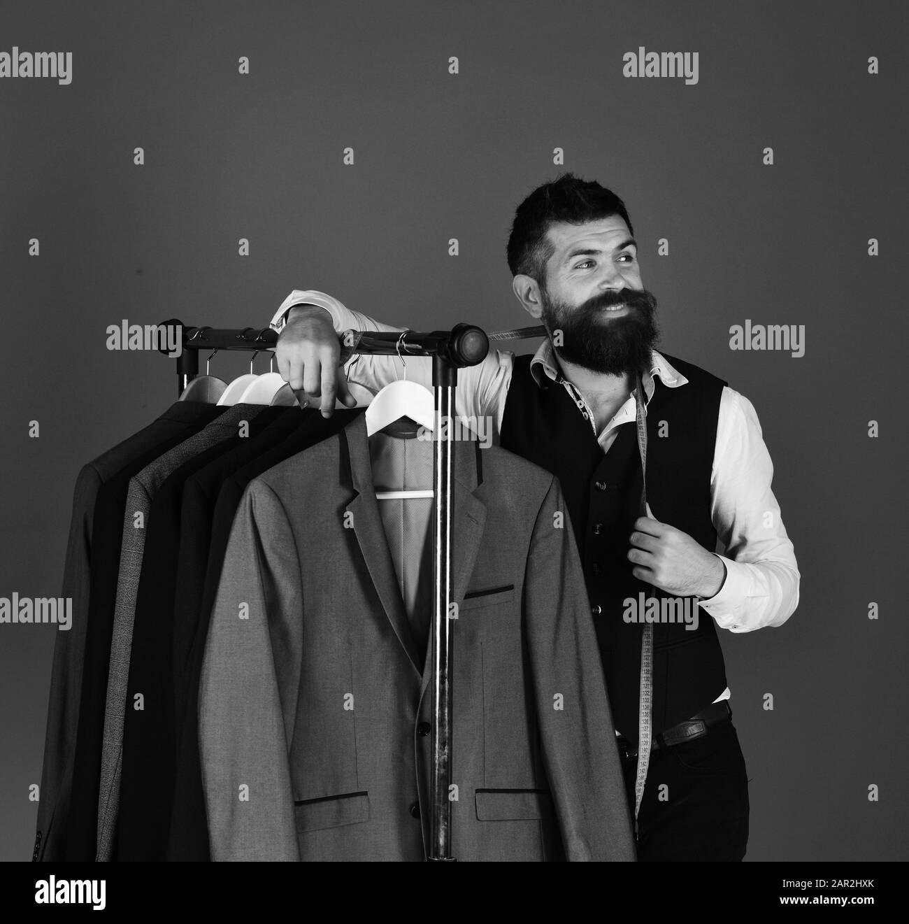 Young tailor with tape measure near suit rack. Guy with beard and ...