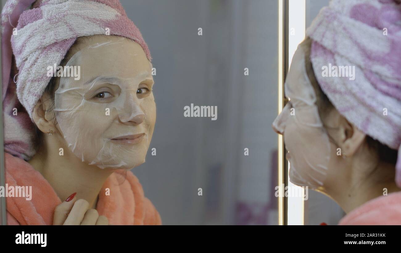 Woman applying cosmetic face mask and looking at mirror in bathroom ...