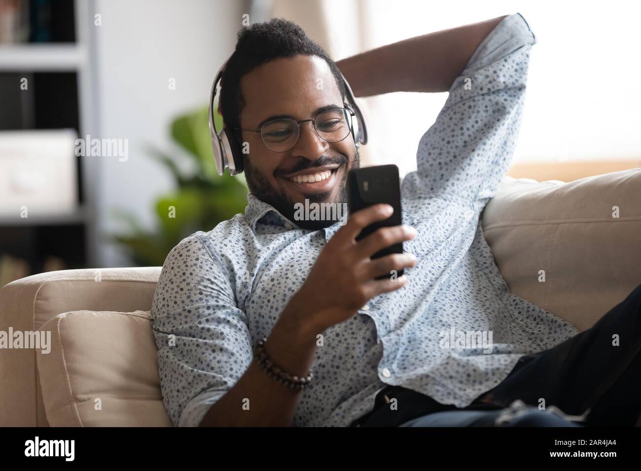 Happy african man wearing headphones listening mobile music on couch ...