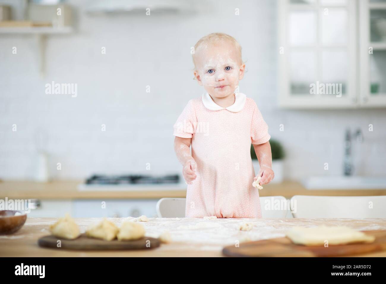 blue-eyed girl with a face in flour holds a piece of dough in her hand ...