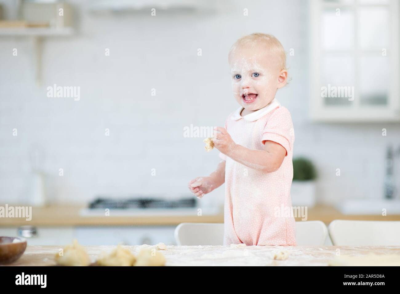 blue-eyed baby girl with a face in flour holds a piece of dough in her ...