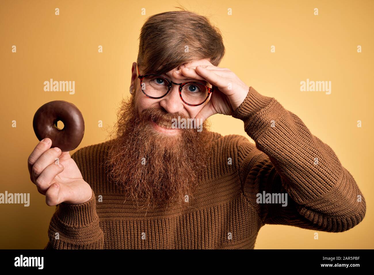 Redhead Irish man with beard eating chocolate donut over yellow ...
