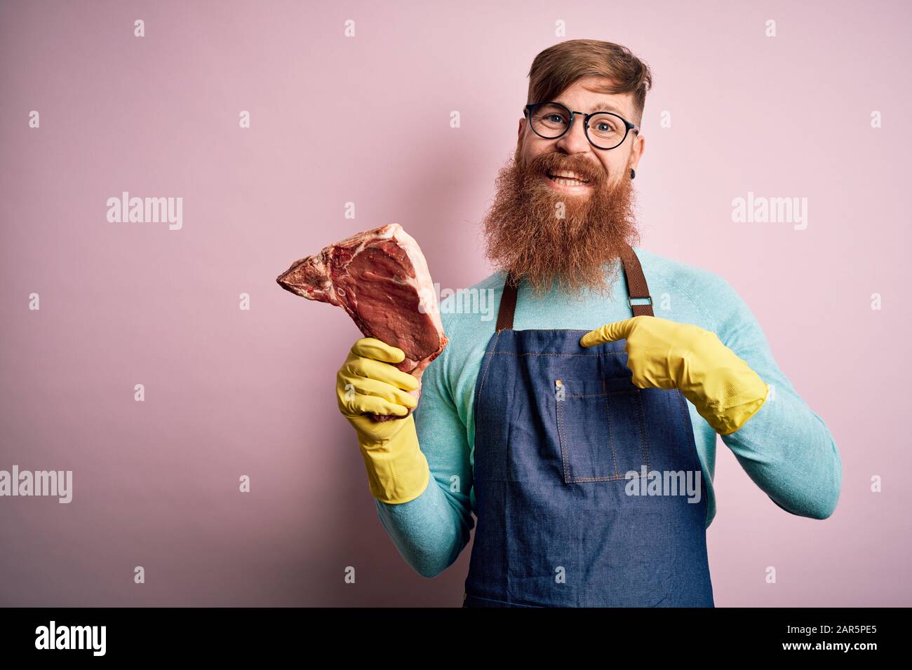 Redhead Irish butcher man with beard holding raw beef steak over pink ...