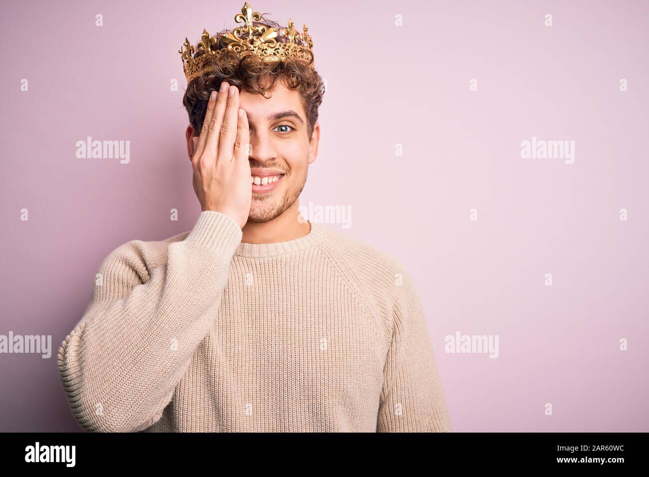 Young blond man with curly hair wearing golden crown of king over pink ...