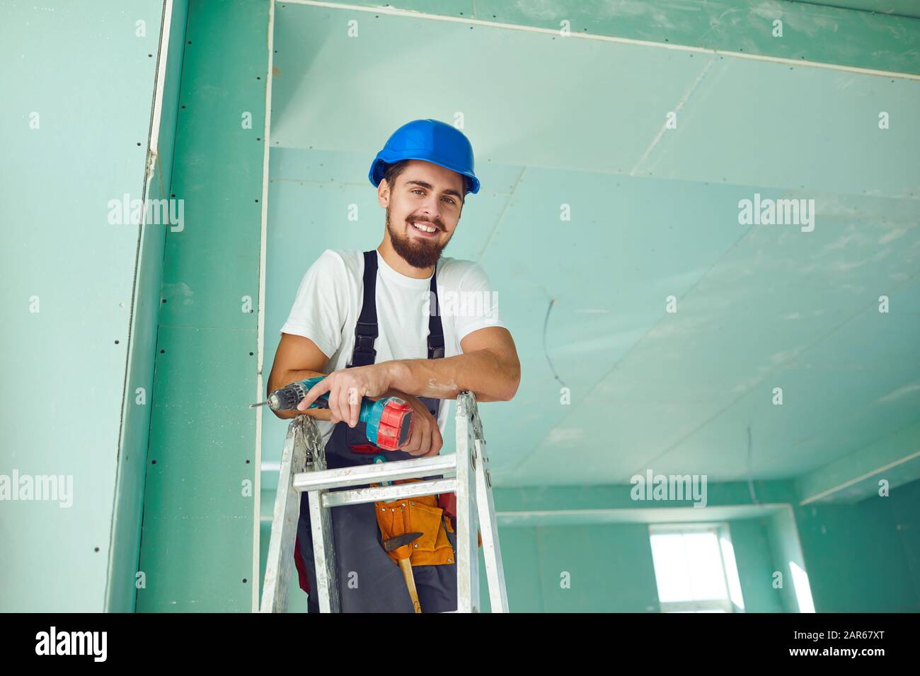 A builder standing on a ladder installs drywall at a construction site ...