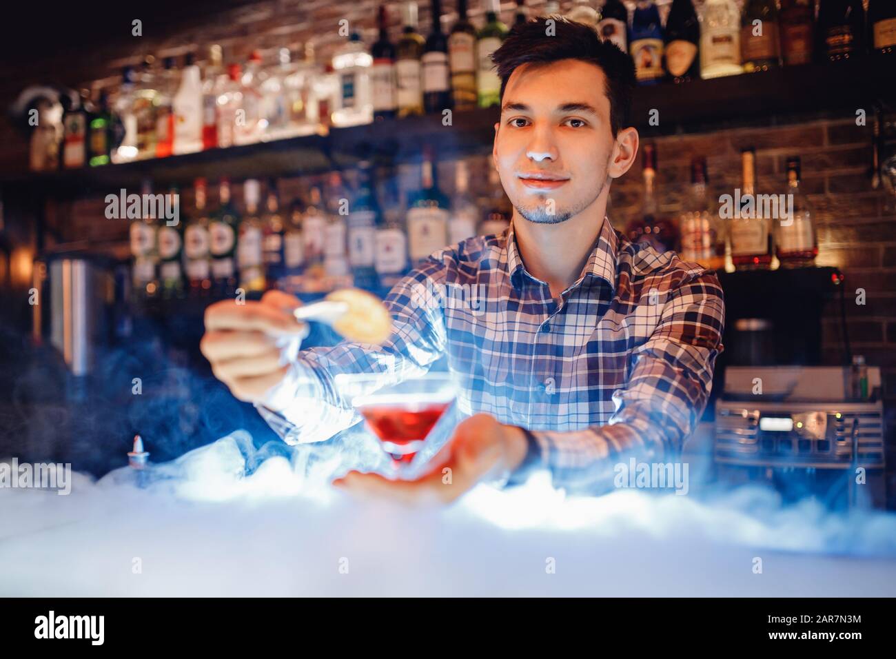 Barman is preparing red alcoholic cocktail with blue smoke in martini ...