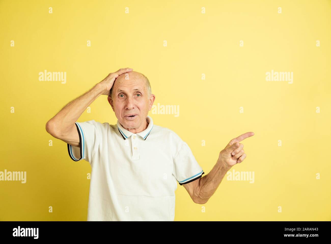 Shocked, pointing at side. Caucasian senior man's portrait on yellow ...
