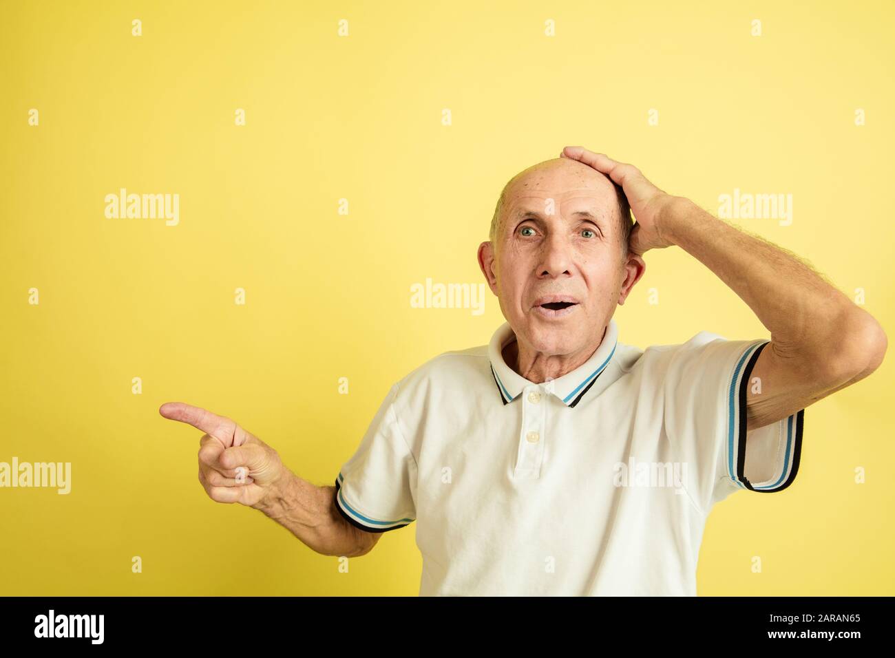 Shocked, pointing at side. Caucasian senior man's portrait on yellow ...