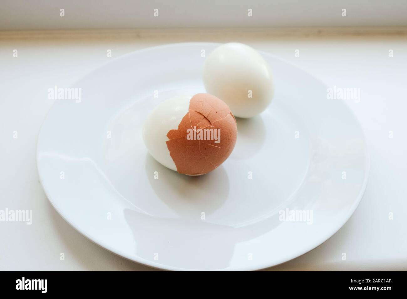 Two boiled eggs on a white plate on a white background. One egg peeled ...