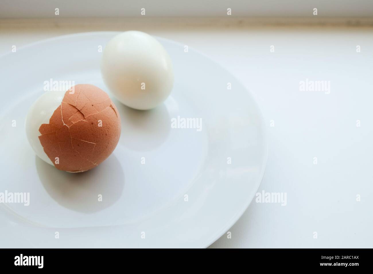Two boiled eggs on a white plate on a white background. One egg peeled ...