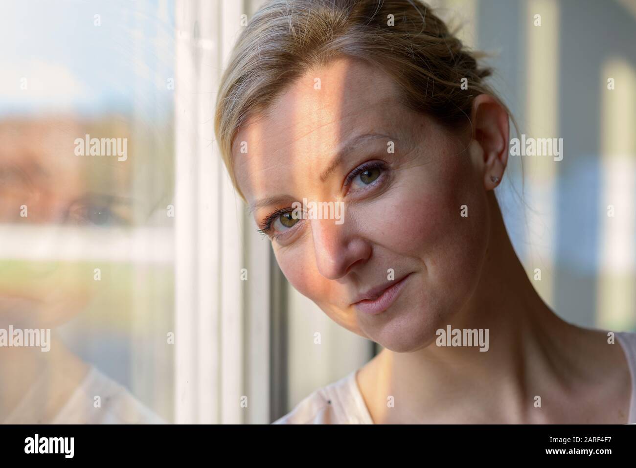 Quizzical blond woman with a knowing smile standing near a window with ...