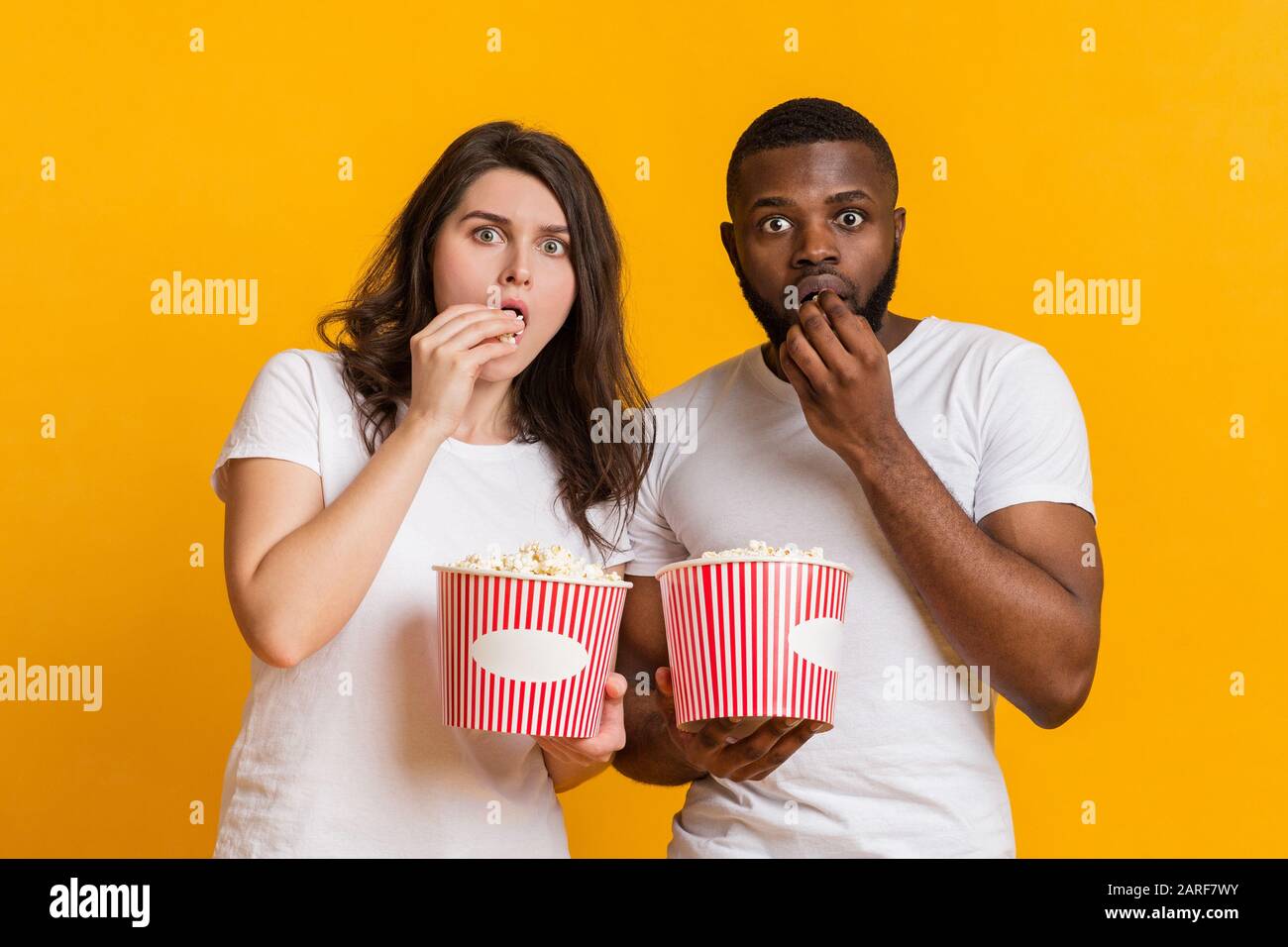 Portrait of dazed interracial couple eating popcorn with scared face ...