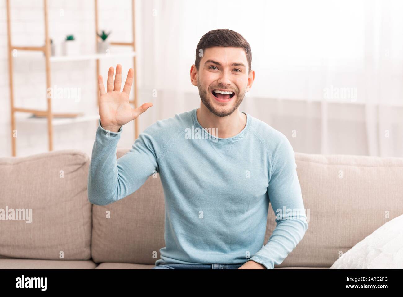 Cheerful Guy Waving Hand Sitting On Couch Indoor Stock Photo - Alamy