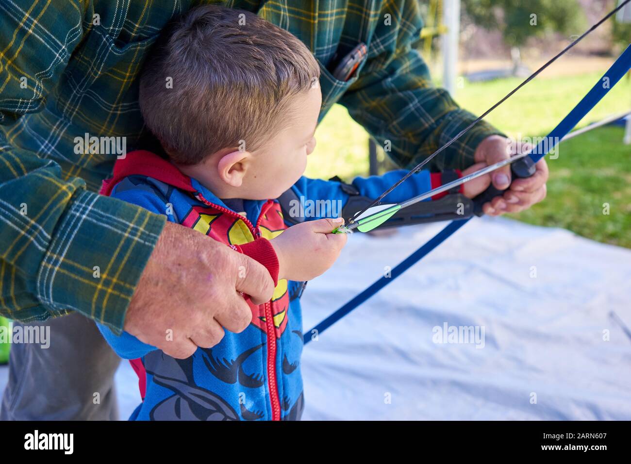 A boy in Spiderman outfit holds bow and arrow as an adult man teaches ...