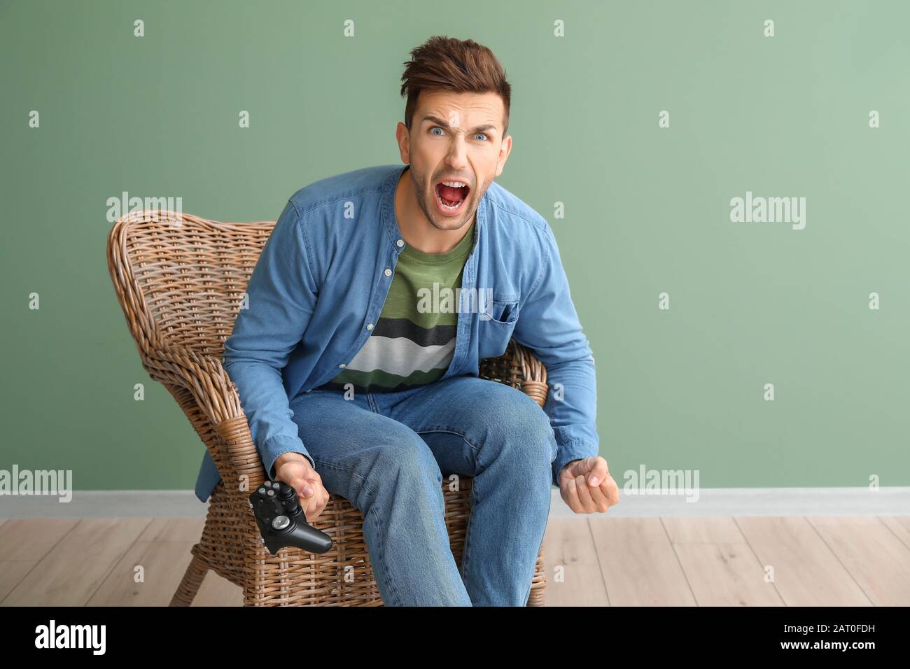Angry man playing video games while sitting in armchair near color wall ...