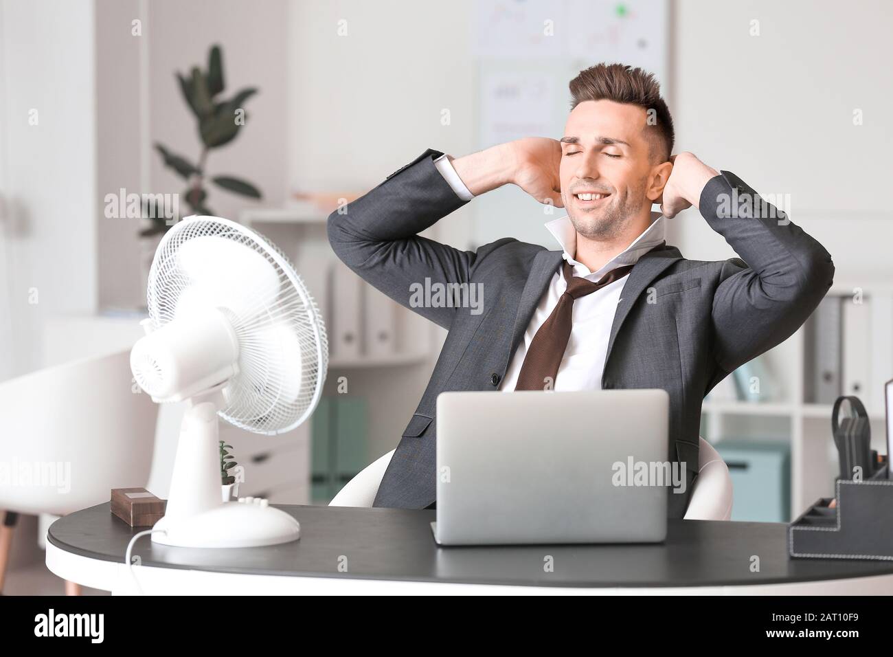 Young man using electric fan during heatwave in office Stock Photo - Alamy