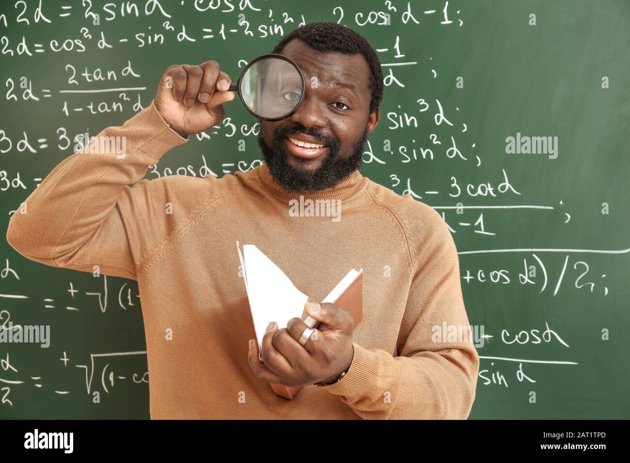 African-American math teacher with book and magnifier near blackboard ...