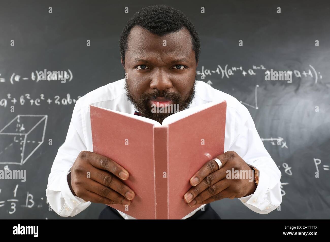 African-American math teacher with book near blackboard in classroom ...