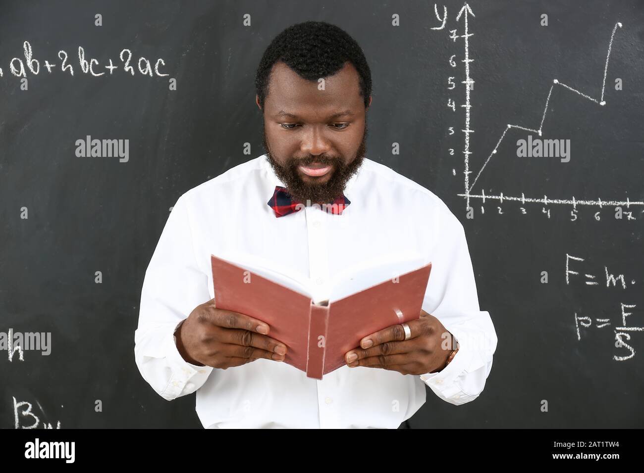 African-American math teacher with book near blackboard in classroom ...