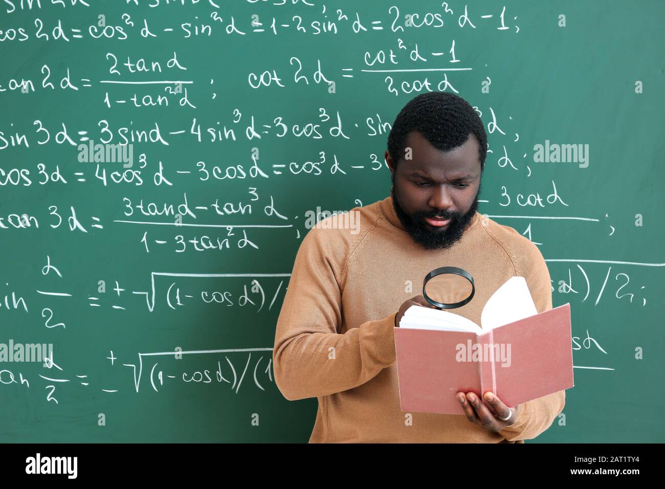 African-American math teacher with book and magnifier near blackboard ...