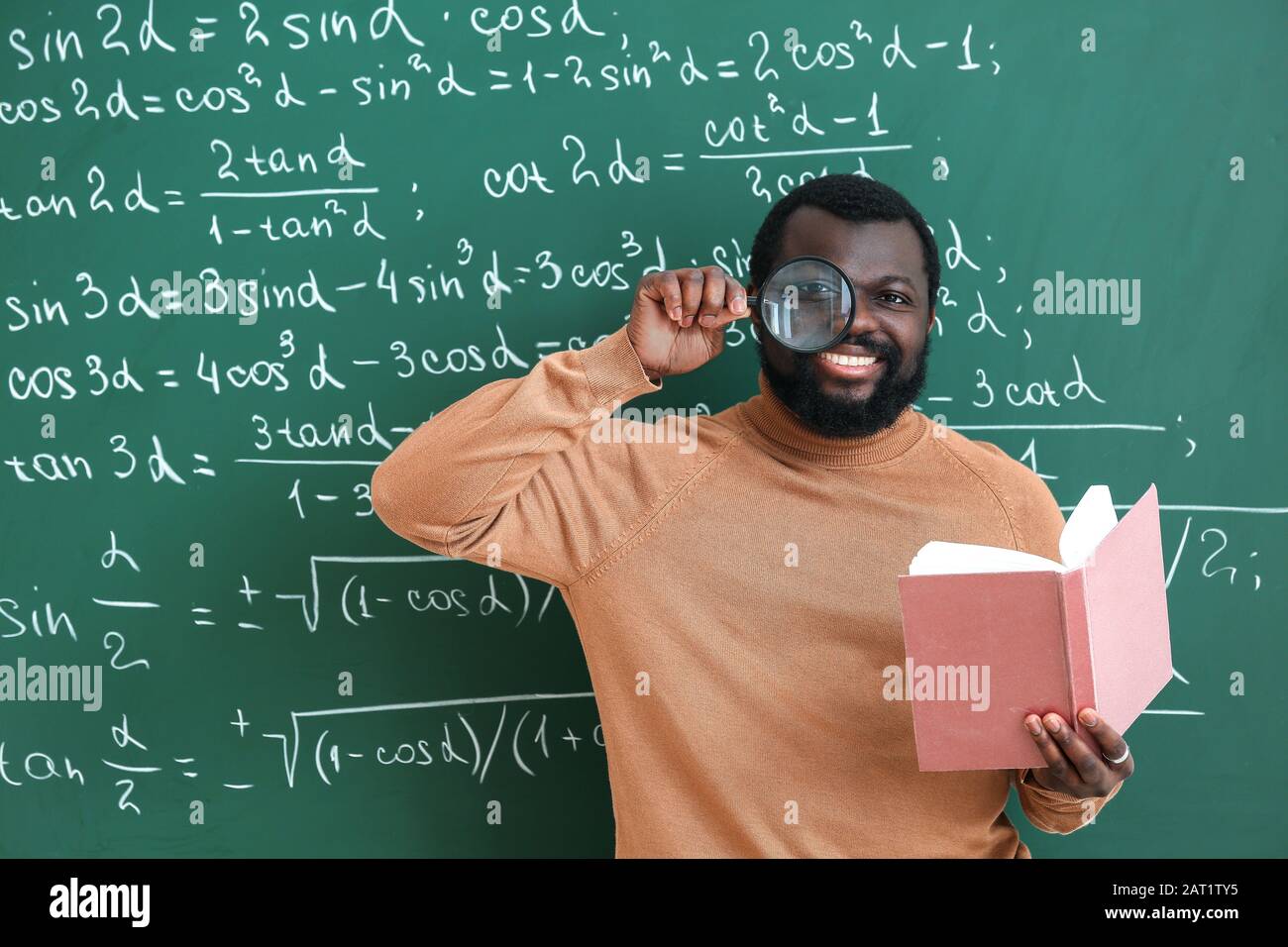 African-American math teacher with book and magnifier near blackboard ...