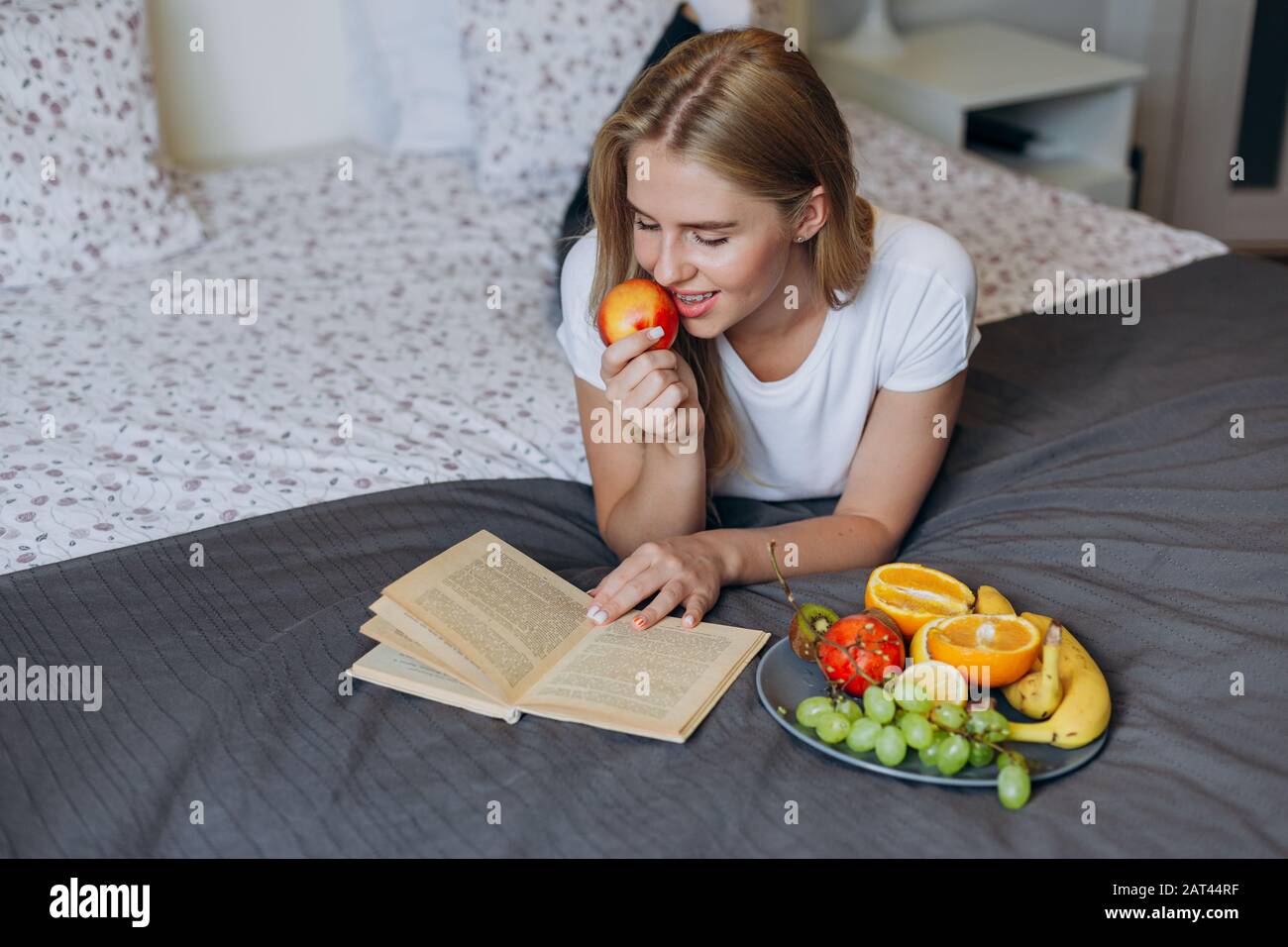 Woman reading a book lying on the bed and eating a healthy food fruits ...