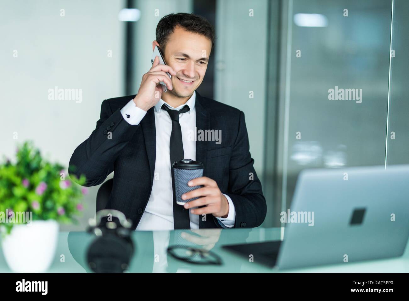 Smiling man talking on mobile phone while using laptop computer at desk ...