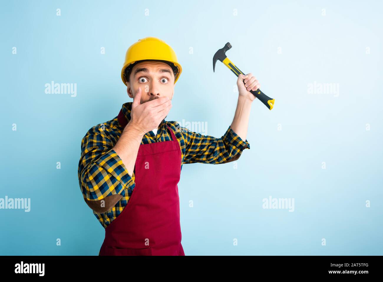 shocked workman holding hammer and covering mouth on blue Stock Photo ...