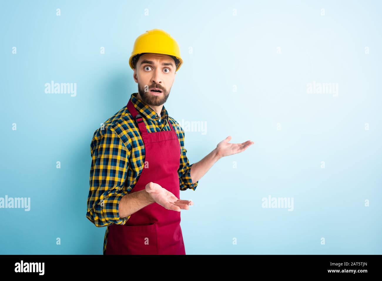 confused workman in safety helmet showing shrug gesture on blue Stock ...