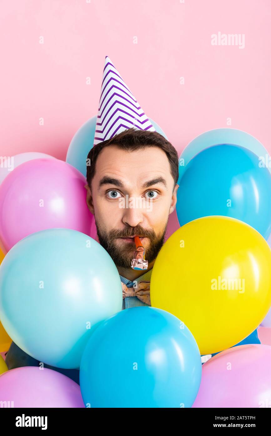 bearded man in party cap blowing party blower near colorful balloons ...