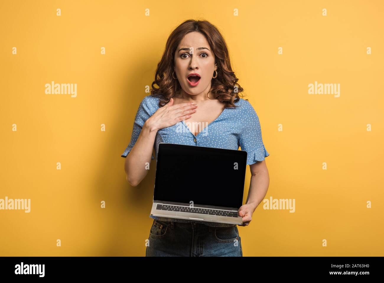 shocked woman touching chest while holding laptop with blank screen on ...