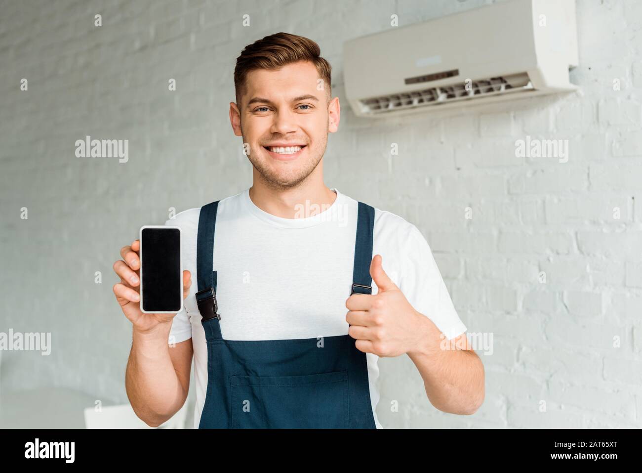 cheerful installer showing thumb up while holding smartphone with blank ...