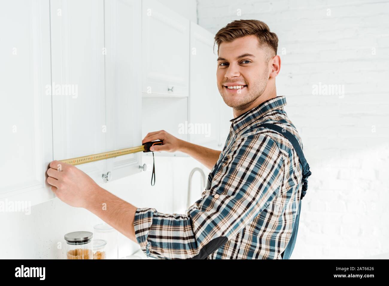 cheerful installer in overalls measuring white kitchen cabinet door ...