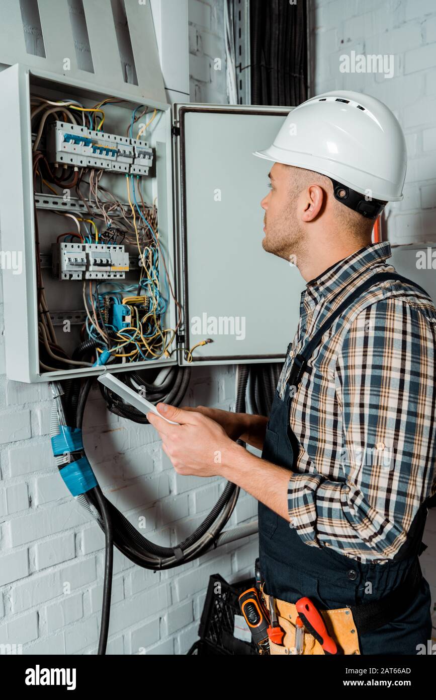 electrician in safety helmet looking at switchboard and holding digital ...